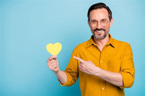 쾌활한 긍정적 인 남자의 초상화 는 그가 14 월 데이트 축하에 그의 아내에서 얻을 노란색 작은 종이 카드 마음을 잡고 파란색 배경 위에 고립 된 캐주얼 스타일의 옷을 입고