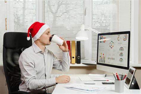 잘생긴 심각한 비즈니스 남자 셔츠와 책상에 앉아 빨간 크리스마스 모자에 흰색 컵에서 차 또는 커피를 마시는 빛 사무실에서 문서와 현대 모니터 컴퓨터에서 작동 경영자에 대한