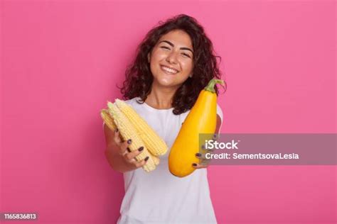 카메라에 애호박과 옥수수를 보여주는 흰색 캐주얼 티셔츠를 입은 젊은 여성의 자른 이미지는 행복한 얼굴 을 발산하고 이빨이 있는 미소로 포즈를 취하고 있습니다 원시 음식