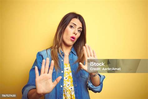 노란색 고립 된 배경 위에 서있는 젊은 아름다운 여성은 두려움과 역겨운 표정으로 거절과 거부를 보여주는 손바닥을 멀리 이동합니다 중지하고 금지 갈색 머리에 대한 스톡 사진