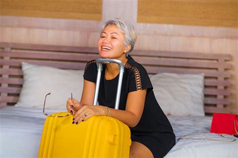 행복 하 고 매력적인 40 대 50 대 성숙한 아시아 관광 여자와 회색 머리에 도착 호텔 객실 흥분 웃는 명랑 즐기는 비즈니스 또는 휴가 여행 편안 하 게 침대에 앉아 여행