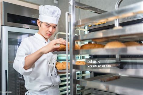 카메라를 보며 웃는 빵 굽는 사람 요리사 드레스와 모자를 쓰고 부엌에서 함께 요리하는 요리사 베이커 젊은 아시아 남자는 부엌에있는 현대 전기 오븐에서 신선한 구운 쿠키를
