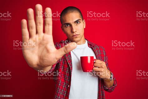 진지하고 자신감있는 표정으로 정지 표지판을하고 열린 손으로 빨간 고립 된 배경 위에 커피 한 잔을 마시는 셔츠를 입은 젊은