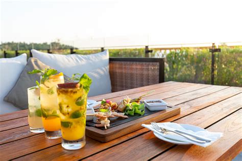 맑은 하늘과 일몰 시간에 잔디 필드 배경이있는 야외 호텔 레스토랑에서 소파와 베개 좌석이있는 나무 테이블에서 저녁 식사 텍스트 공간을 복사 칵테일에 대한 스톡 사진 및 기타