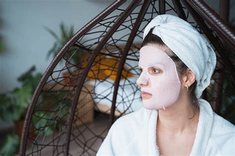 아름다운 젊은 여성은 시트 보습 얼굴 마스크를 사용합니다 녹색 식물아파트에 나무 스윙 의자를 매달려 화이트 목욕 가운 수건 셀프 스킨 케어 집에서 주름에 대한 안티 에이지