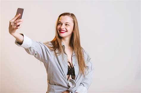 가벼운 캐주얼 의류의 행복 한 귀여운 소녀는 셀 카를 만들고 스마트 폰을 살펴보고 미소를 짓습니다 귀여운 금발 머리와 빨간 입술 여자는 전화에 사진을 만든다 감정에 대한