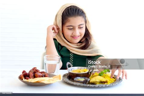 부엌 식탁에 앉아 아름다운 눈을 가진 사랑스러운 웃는 파키스탄 무슬림 소녀 히잡과 전통 의상을 입은 아이는 하얀 배경에 손으로 전통적인 이슬람 할랄 음식을 먹는 것을 즐긴다