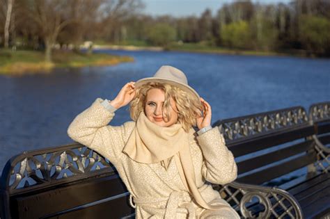 강가에서 베이지색 코트를 입은 여자가 미소를 지으며 봄 풍경을 배경으로 바람에 맞서 모자를 들고 있다 30 34세에 대한 스톡 사진 및 기타 이미지 Istock