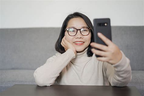 스마트 폰을 들고 있는 젊은 아시아 여성이 주말에 집 안의 거실에서 친구 및 가족과 소통하여 사회적 거리와 격리 시간 개념을 확보했습니다 40 44세에 대한 스톡 사진 및