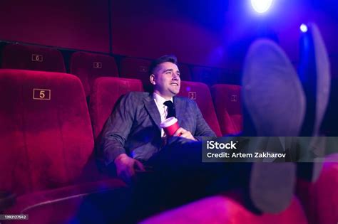 비즈니스 슈트를 입고 영화관에서 홀로 젊고 잘 생긴 남자의 초상화 앞 좌석에 발을 얹고 빨간 컵에서 튜브를 통해 술을 마시는 모습 Premiere에 대한 스톡 사진 및 기타