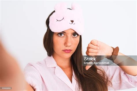 잠옷과 수면 마스크를 착용한 소녀는 화가 난 얼굴로 고립 된 흰색 배경 위에 카메라로 셀카를 만들고 엄지 손가락으로 싫어하는