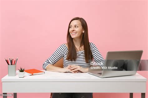 캐주얼 옷 옆으로 보이는 젊은 여 자가 앉아 파스텔 핑크 배경에 고립의 현대 Pc 노트북과 흰색 책상에서 일 성과 비즈니스 경력 개념입니다 광고에 대 한 공간을 복사 합니다