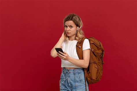 빨간 배경에 서 흰색 티셔츠를 입고 곱슬 금발 머리를 가진 여자의 초상화 주황색 배낭 흰색 헤드셋 및 휴대 전화가 카메라를 보고 충격을 받은 모델 T 셔츠에 대한 스톡 사진