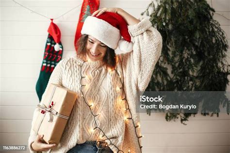 현대 크리스마스 트리 조명및 스타킹의 배경에 크리스마스 선물 상자를 들고 산타 모자와 아늑한 스웨터에 세련된 행복 소녀 젊은 힙스터 여자 들고 선물 과 미소 12월에 대한