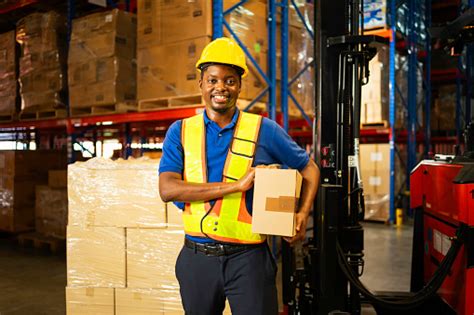 안전 장비를 착용한 미소 짓는 창고 직원이 바쁜 산업 환경에서 효율성과 팀워크를 상징하는 상자를 들고 있습니다 35 39세에 대한 스톡 사진 및 기타 이미지 Istock