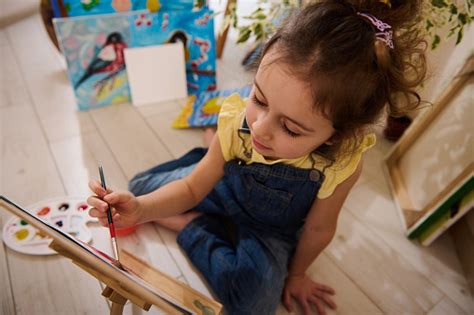 노란 티셔츠와 파란색 데님 페인팅을 입은 클로즈업 어린 소녀가 집에서 아름다운 그림을 그린 나무 이젤 옆에 있는 바닥에 앉아 있습니다 어린이 예술과 어린이 취미 교육 개념