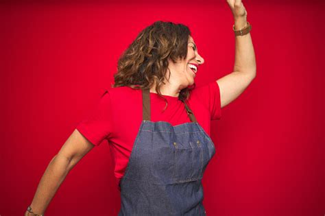 빨간 고립 된 배경에 앞치마 유니폼을 입고 중년 시니어 여성은 행복하고 쾌활한 춤을 추며 캐주얼하고 자신감있는 음악을 듣고 미소짓습니다 앞치마에 대한 스톡 사진 및 기타