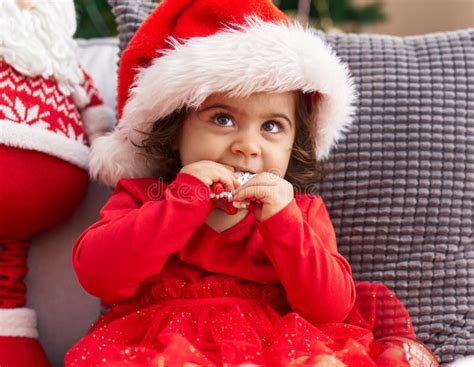 Adorable Hispanic Girl Sucking Stair Decoration Sitting On Sofa By Christmas Tree At Home Stock