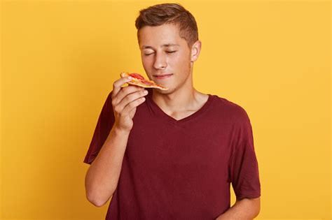 즐거운 달콤한 어린 소년의 초상화는 그의 눈을 감고 피자 조각을 들고 피자를 좋아하고 캐주얼 한 어두운 빨간 티셔츠를 입고 스튜디오에서 노란색 배경 위에 고립 되어 있습니다