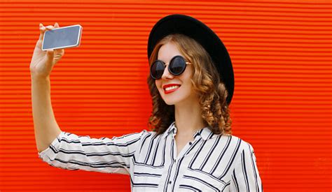 흰색 줄무늬 셔츠 빨간 배경 위에 검은 둥근 모자를 입고 전화로 셀카 사진을 찍는 행복한 미소 여자의 초상화 20 29세에 대한