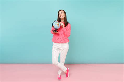 니트 장미 스웨터 흰색 바지 스튜디오에서 분홍색 파란색 파스텔 벽 배경에 고립 된 지구 세계 글로브와 포즈의 여자의 전체 길이 초상화 패션 라이프 스타일 개념입니다 복사