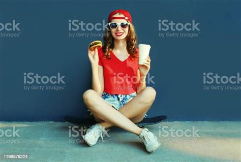야구 모자를 쓰고 스케이트 보드에 앉아 햄버거와 커피 컵시원한 젊은 여자 푸른 벽 배경 위에 선글라스 거리에 대한 스톡 사진 및