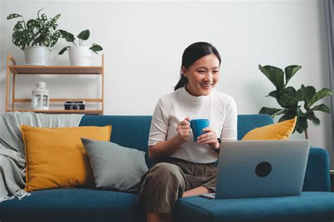 아시아 여성들은 커피잔을 들고 노트북을 사용하여 집 거실에 앉아 일하거나 인터넷 서핑을 한다 여자에 대한 스톡 사진 및 기타 이미지 Istock