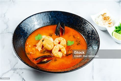 해산물과 베트남 어 톰 얌 수프 새우 가리비 홍합 연어를 대리석 테이블에 어두운 세련된 그릇에 담은 붉은 수프 미니멀 한 음식 사진 개념입니다 부야베스 0명에 대한 스톡