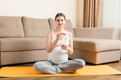 요가 매트에 연꽃 위치에 앉아있는 동안 물 병을 가진 임신 한 젊은 여성 코로나바이러스 시간에서 물 균형 유지 임신에 대한 스톡