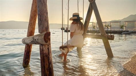 긴 흰색 드레스를 입은 젊은 아름 다운 여성이 일몰 광선이 비추는 바다 해변에서 밧줄 그네에서 휴식을 취하고 있습니다 관광