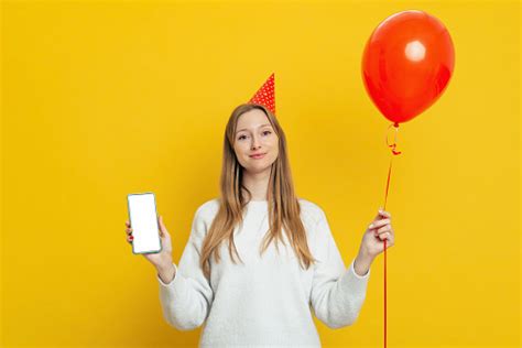 광고를위한 빈 화면이있는 휴대 전화를 들고 흰색 캐주얼 스타일 스웨터를 착용하고 빨간색 풍선으로 노란색 배경 위에 서있는 축하 모자에 행복한 미소 짓는 젊은 여성 25 29