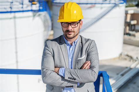 팔을 교차하고 카메라를 보고 높이에 서서 머리에 헬멧을 쓰고 양복을 입은 매력적인 면도하지 않은 사업가 정유 공장 외관 배경에는