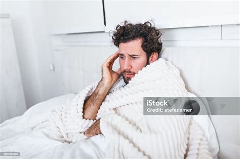 담요 아래에 앉아 두통을 가진 아픈 사람 계절 감염 독감 침대에 누워 알레르기가있는 아픈 사람 아픈 잘생긴 남자는 고열과 독감 휴식과 침대에 누워 담요로 덮여 재채기에 대한