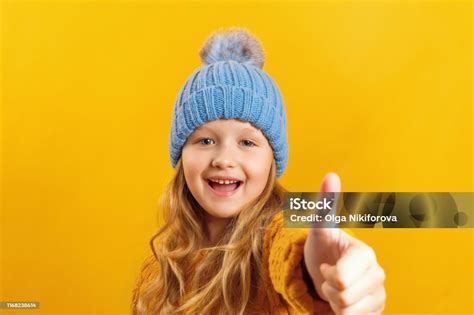 스웨터와 모자를 쓰고 있는 쾌활한 어린 소녀 엄지손가락을 치켜들고 노란 배경에 카메라를 바라보고 있습니다 가을에 대한 스톡 사진 및 기타 이미지 Istock