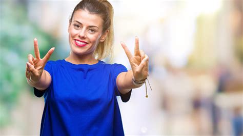 손가락 하 고 승리 기호를 보여주는 카메라를 보고 웃 고 고립 된 배경 위에 캐주얼 블루 티셔츠를 입고 젊은 아름 다운 여자