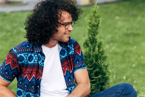 곱슬 머리를 웃고 화려한 셔츠와 공원벤치에 앉아 투명 안경을 입고 백인 젊은이 화창한 여름날을 즐기는 캐주얼 한 트렌디 한 의상을 입은 쾌활한 남성 20 29세에 대한 스톡