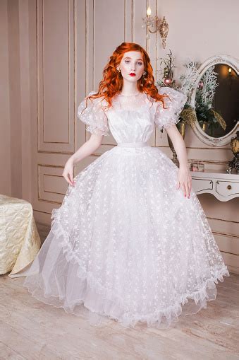 그녀의 귀에 흰색 진주 귀걸이와 흰색 빈티지 웨딩 드레스에 긴 붉은 곱슬 머리를 가진 여자 창백한 피부 파란 눈 고급 스러운 침실에 특이 한 외관을 밝은 빨간 머리 소녀