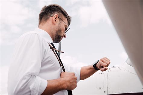 비행기 근처에 서서 손목시계를 보고 있는 선글라스를 착용한 수염 조종사의 낮은 각도 보기 낮은 카메라 각도에 대한 스톡 사진 및