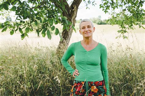 녹색 셔츠 화려한 꽃 바지 호두 나무 앞에서 녹색 초원에서 포즈를 취하는 짧은 회색 머리카락을 가진 예쁜 중년 여성 50 54세에 대한 스톡 사진 및 기타 이미지 Istock