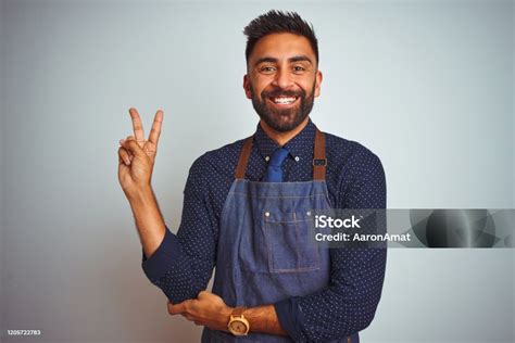 고립 된 흰색 배경 위에 서있는 앞치마 유니폼을 입은 젊은 인도 직원 남자가 승리의 표지판을하고있는 카메라에서 행복한 얼굴 윙크와 미소 2번 몸짓에 대한 스톡 사진 및 기타