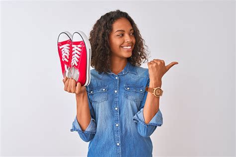 고립된 흰색 배경 위에 서있는 운동화를 들고 있는 젊은 브라질 여성이 행복한 얼굴미소를 지으며 엄지손가락을 들고 옆으로 미소 짓고 있는 모습 가리키기에 대한 스톡 사진 및