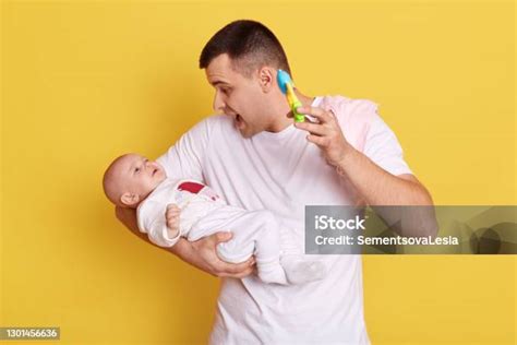 딸랑이 장난감을 들고 놀고 노란 배경위에 고립 된 아기를 진정시키고 아빠가 노래를 부르거나 손에 들고 아이를 위해 무언가를 소리치고 남성은 흰색 캐주얼 티셔츠를 입고