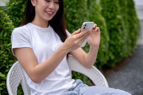 A Positive Asian Woman Is Scrolling On Her Phone While Relaxing Sitting On A Chair In A Garden