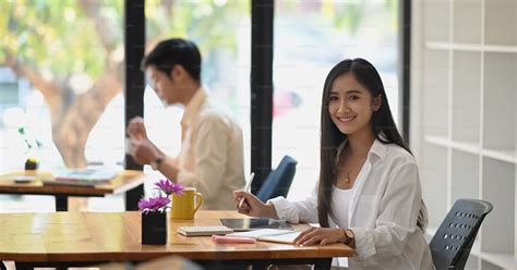 흰색 셔츠를 입은 젊은 아름다운 여성이 스타일러스 펜으로 컴퓨터 태블릿에 그림을 그리는 동안 동료와 현대적인 사무실을 배경으로 작업 나무 테이블에 앉아 있는 측면 사진