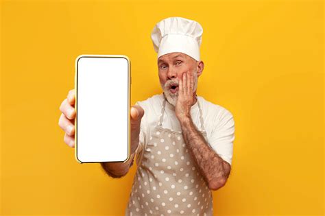 모자와 앞치마를 입은 늙은 대머리 할아버지 요리사가 노란색 고립 배경에 빈 스마트폰 화면을 보여줍니다 70 79세에 대한 스톡 사진 및 기타 이미지 Istock