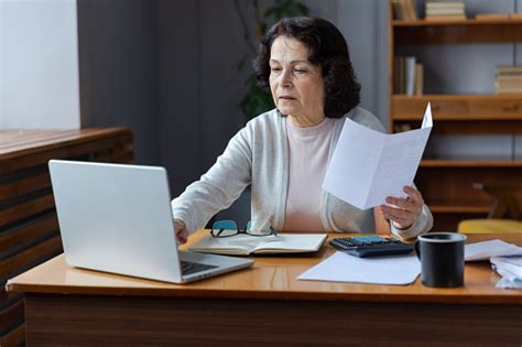 중년의 고위 여자는 노트북과 종이 문서 잠겨 있는 나이 든 성숙한 여자는 종이 청구서를 읽고 집에서 온라인으로 지불 은행 재정 계산 세금 계획 대출 부채 연금 지불 65