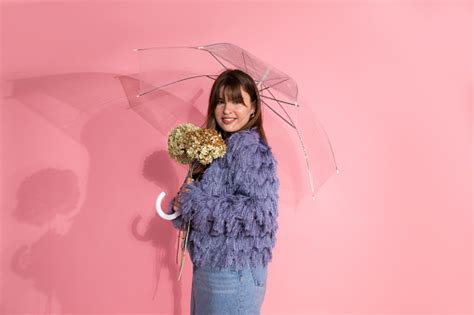 젊고 아름답고 검은 머리의 여성이 투명한 우산 아래 스튜디오의 분홍색 배경에 서서 손에 있는 꽃이 카메라를 바라보며 미소를 짓고 있습니다 꽃 식물에 대한 스톡 사진 및 기타