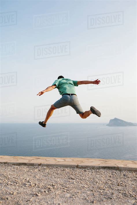 Low Angle Back View Of Unrecognizable Male In Casual Clothes Jumping High Against Blue Sky And