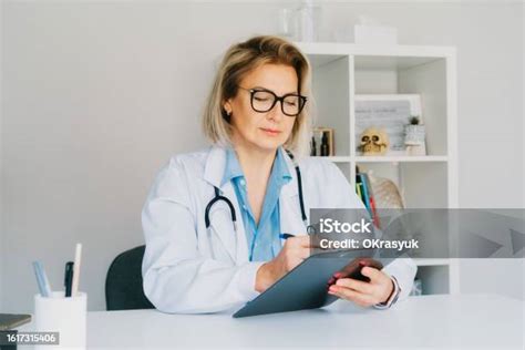 자신감 넘치는 성숙한 중년 여성 의사 의료 간호사 흰 코트를 입은 여성 의사가 청진기를 들고 병원 사무실의 작업장에 앉아 약속을