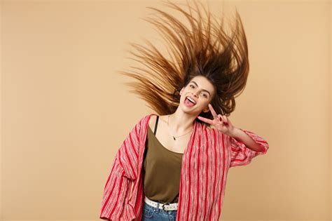 캐주얼에 쾌활 한 젊은 여자의 초상화 파스텔 베이 지 배경에 고립 된 승리 기호를 보여주는 흐르는 머리와 재미 사람들은 진심으로 감정 라이프 스타일 개념입니다 복사 공간을